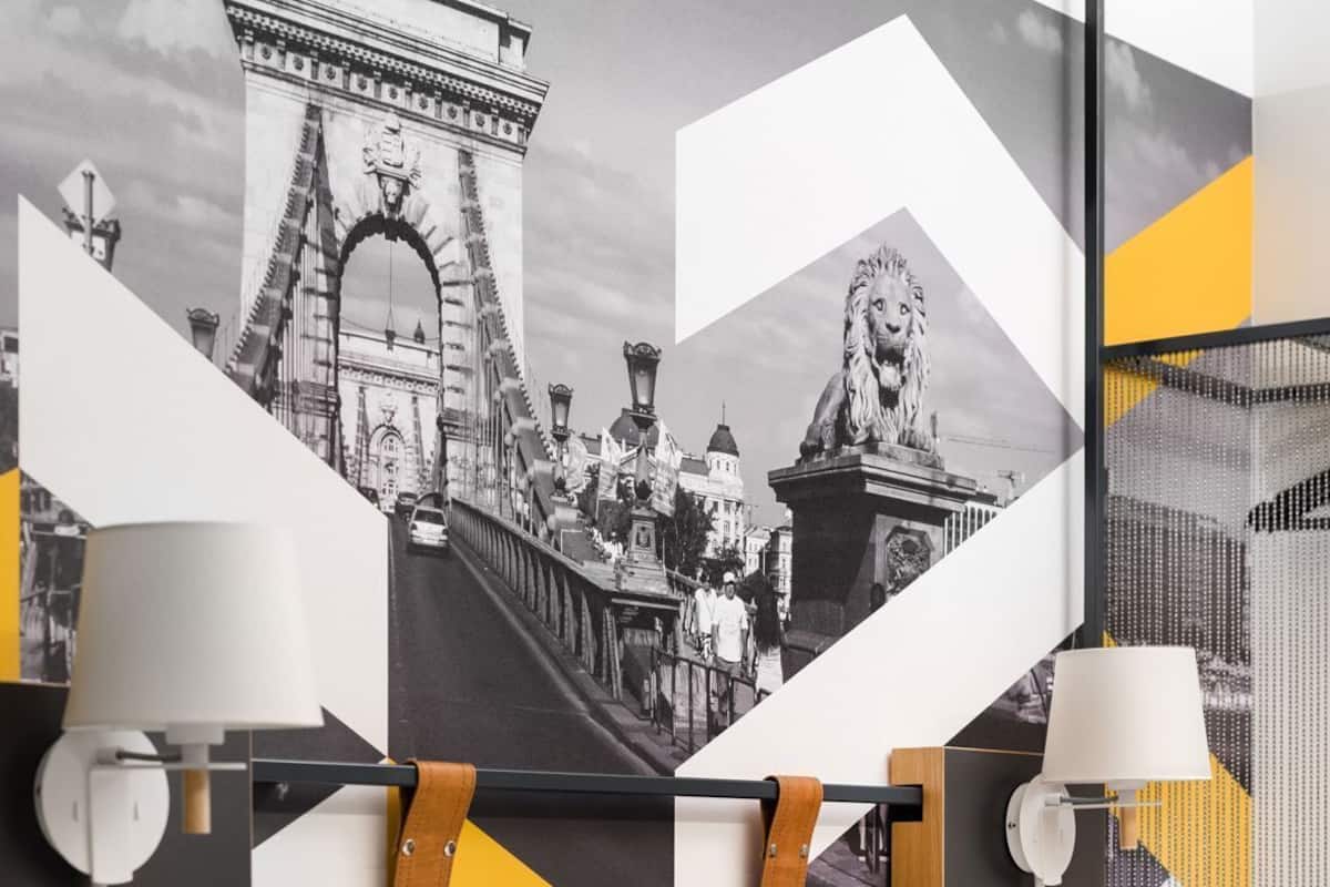 Quarto de hotel com cama, almofadas, abajur, revista e parede com fotografia em preto e branco.