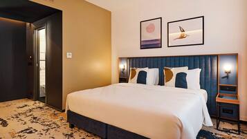 Quarto de hotel com uma cama grande, cabeceira com iluminação embutida, mesa de cabeceira pequena e duas obras de arte penduradas na parede.