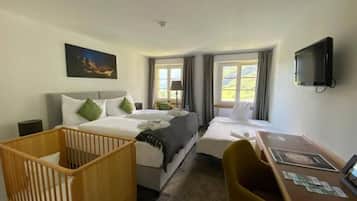 Quarto de hotel com duas camas, um bercinho de madeira, uma escrivaninha com cadeira, televisão e uma janela com cortinas.