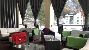 Uma área de estar acolhedora com móveis de vime e uma mesa de centro de vidro, com vista para uma montanha nevada e edifícios.