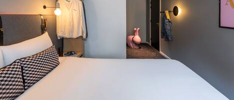 Quarto de hotel moderno com uma cama grande, um roupão pendurado, uma cadeira de coelho rosa peculiar e um quadro na parede.