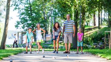 Uma família jogando croquet em um caminho pavimentado em um parque, com árvores e vegetação ao fundo.