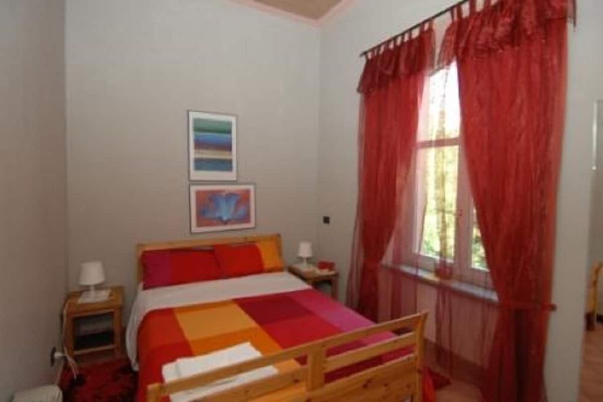 Um quarto com cama, mesa de cabeceira, janela com cortinas vermelhas e duas fotos emolduradas na parede.