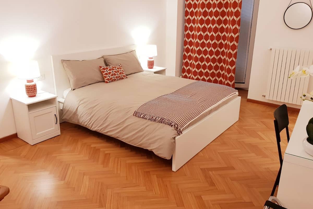 Um quarto bem arrumado com uma cama, mesas de cabeceira, um radiador e uma cadeira.