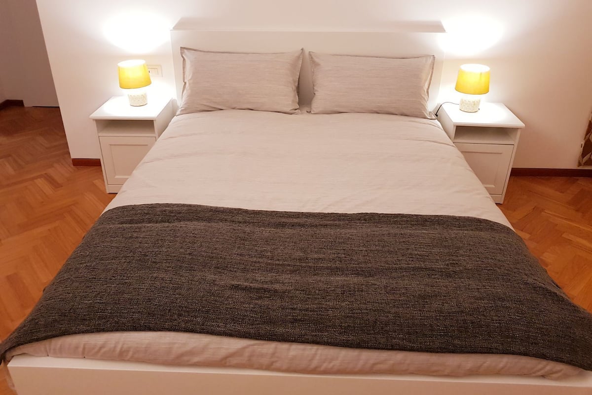 Uma cama com um cobertor cinza, duas luminárias de cabeceira e uma cabeceira branca.
