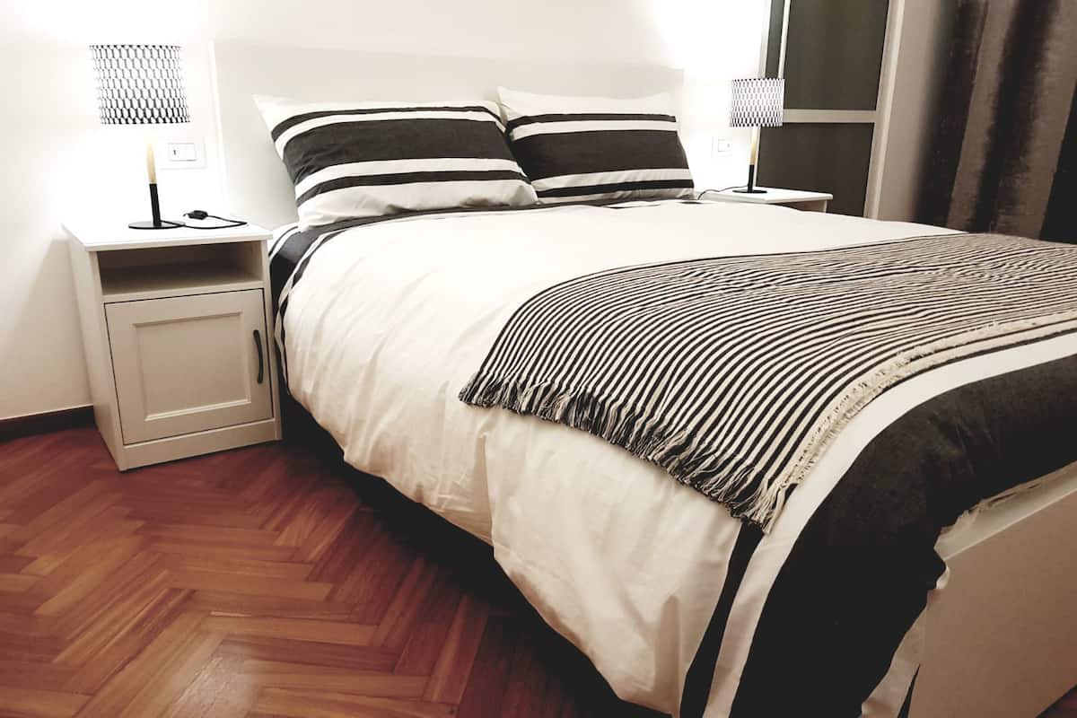 Cama bem arrumada com manta listrada e almofadas, mesa de cabeceira com abajur e piso de madeira.