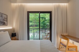 Quarto de hotel com uma cama, uma escrivaninha, uma cadeira, um abajur e vista para o verde através de uma grande janela.