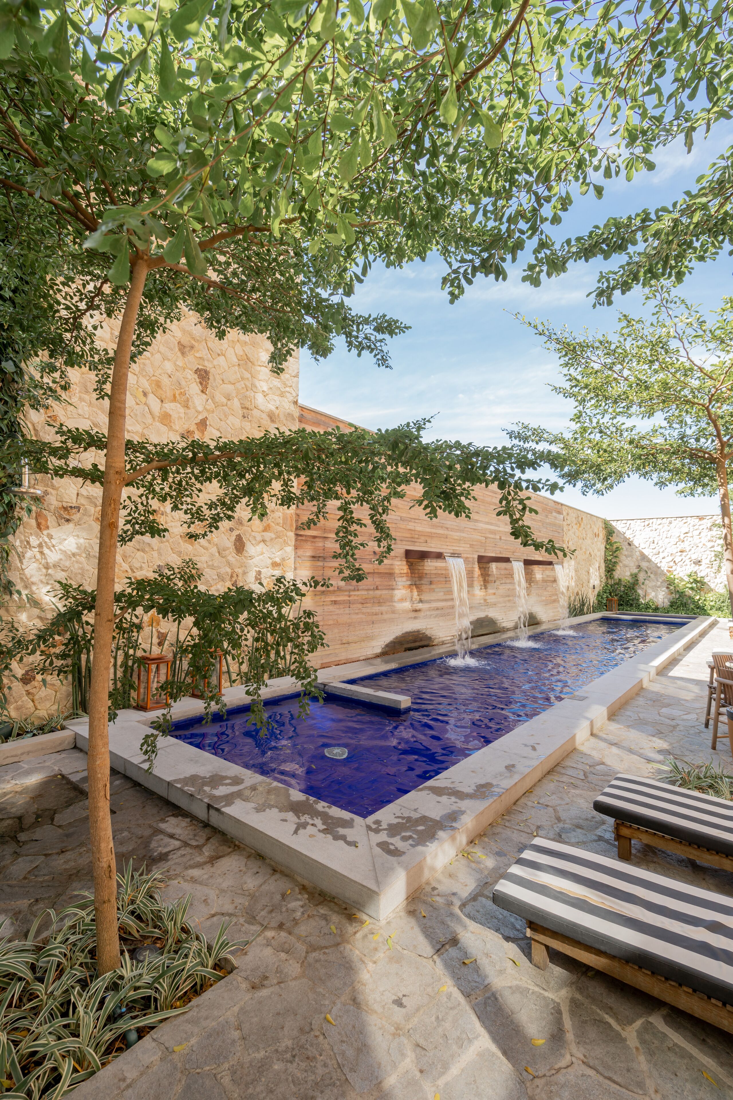 Uma área de piscina com muro de pedra, uma árvore e espreguiçadeiras.
