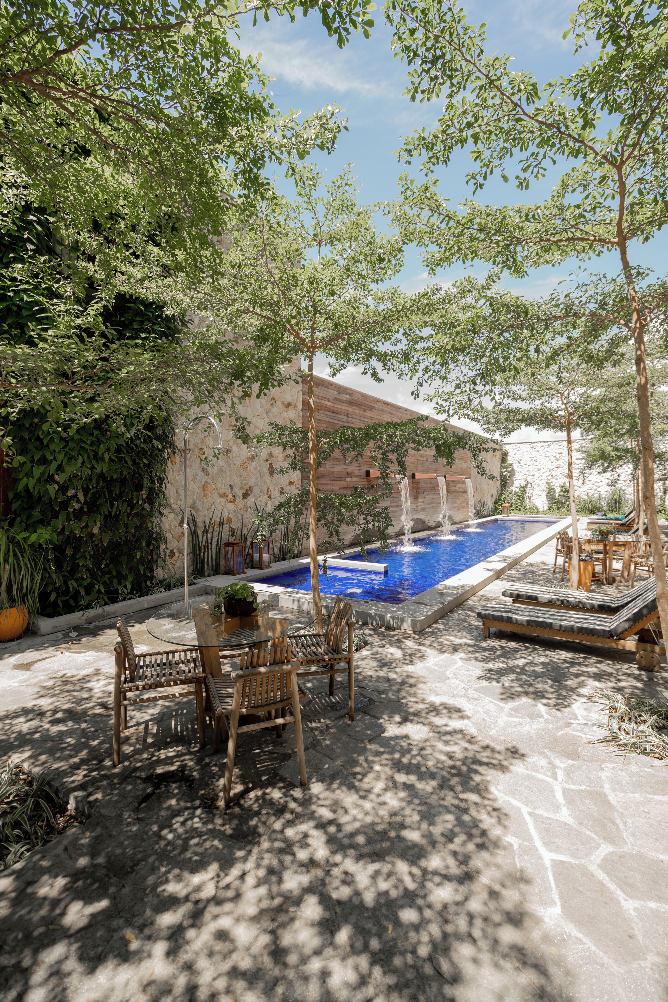Uma área de piscina com mesa e cadeiras, cercada por vegetação e um muro de pedra.