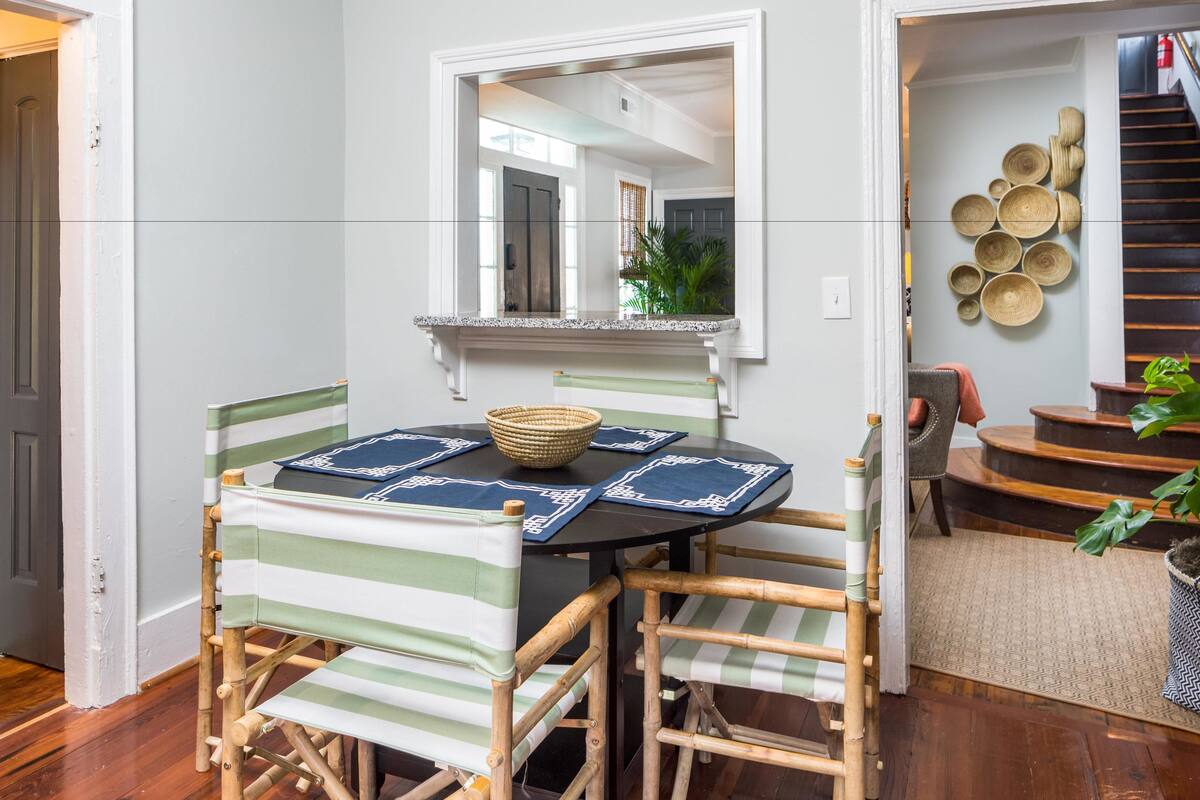 Uma sala de jantar com uma mesa posta para quatro pessoas, rodeada por cadeiras listradas, um espelho acima da mesa e uma escadaria para outro cômodo.