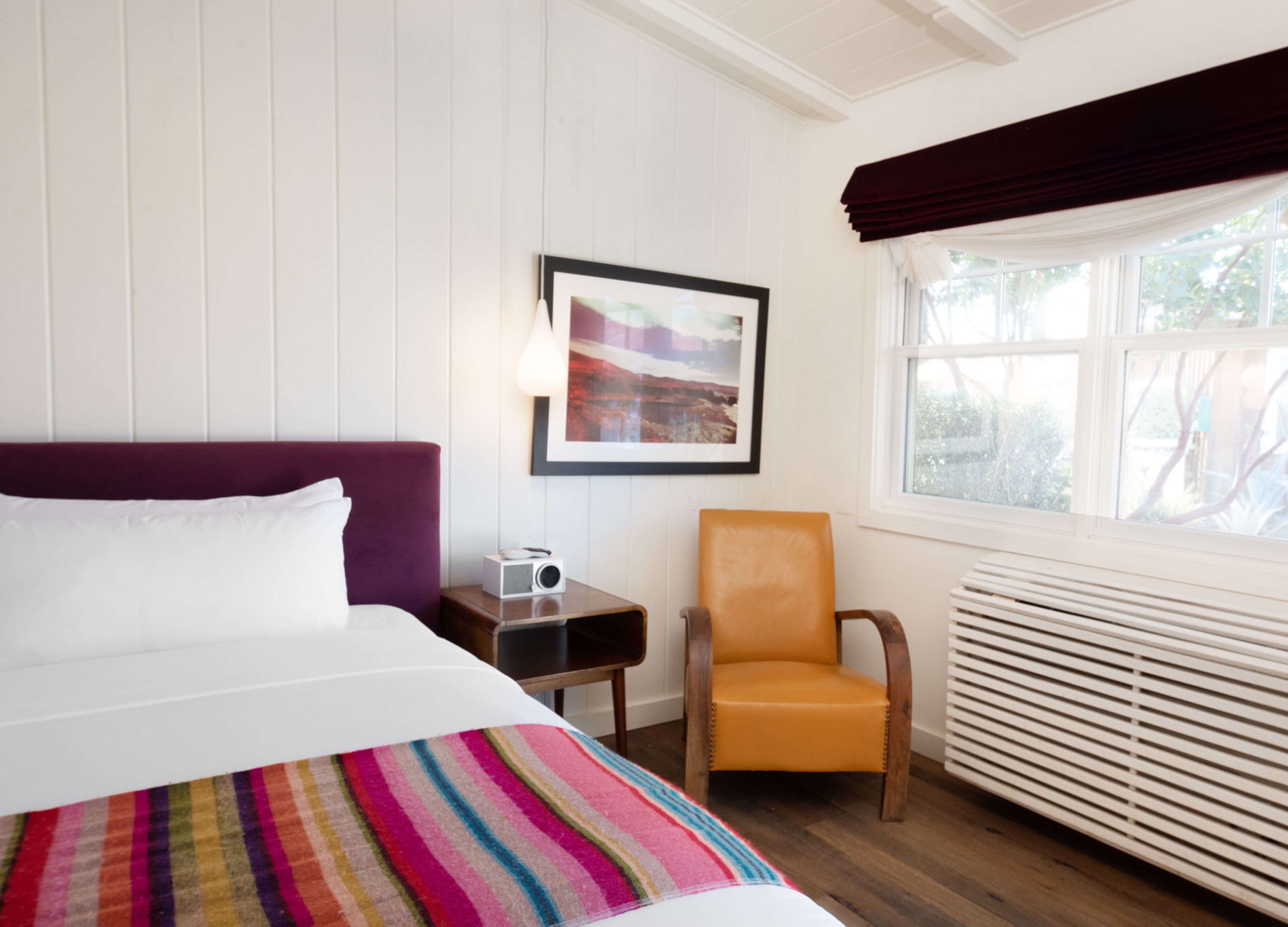 Quarto de hotel com cama, cadeira, mesa de cabeceira, quadro e janela com vista.