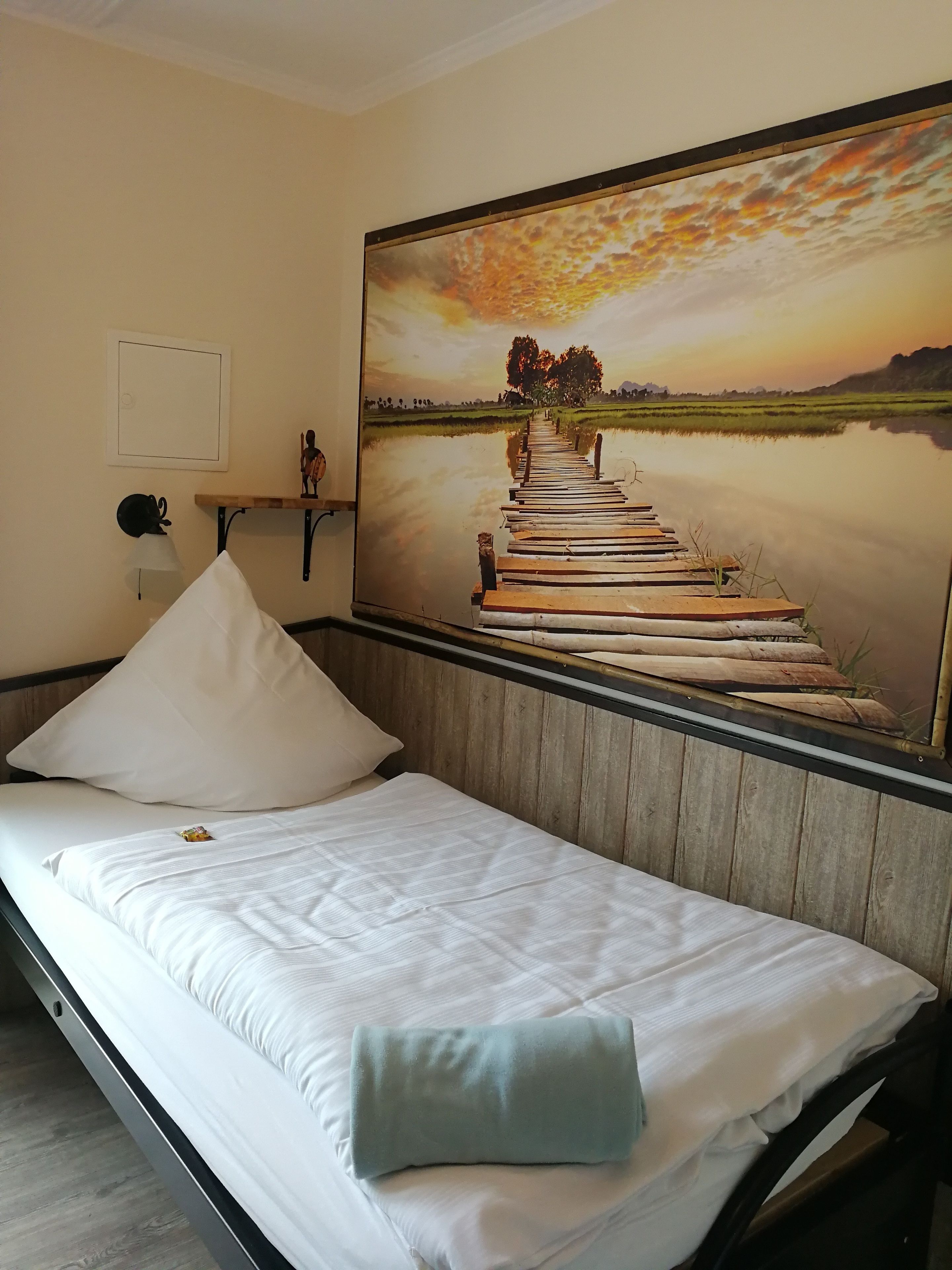 Uma cama bem arrumada com travesseiro e manta dobrada, prateleira na parede com abajur, e uma grande pintura de um pôr do sol sobre um rio.
