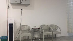 Quarto com uma pequena mesa e três cadeiras de vime, um bebedouro e uma tomada na parede.