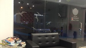 Um hall de hotel moderno com um sofá de couro preto, uma pequena mesa redonda com livros e uma parede de vidro com vista para a cidade.