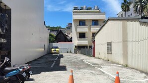 Uma rua urbana estreita com uma moto estacionada e um carro amarelo, dois cones de trânsito laranja, e um edifício com várias sacadas ao fundo.