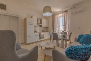 Uma sala de estar moderna com sofás cinza, uma mesa redonda pequena e um espaço de refeições com mesa e cadeiras brancas. Há uma cozinha com armários brancos e um lustre.