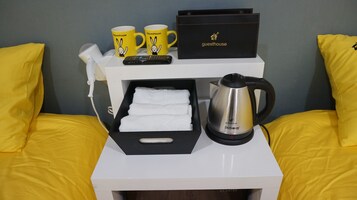 Quarto de hotel bem organizado, com uma cama, uma mesinha de cabeceira, um chaleira, duas canecas, um controle remoto e uma caixa preta com o logo 