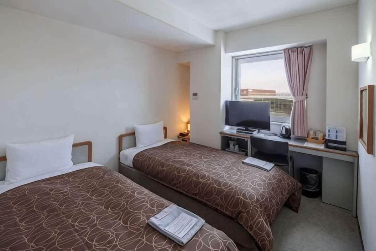 Quarto de hotel com duas camas, uma secretária com computador, televisão e janela com cortinas.