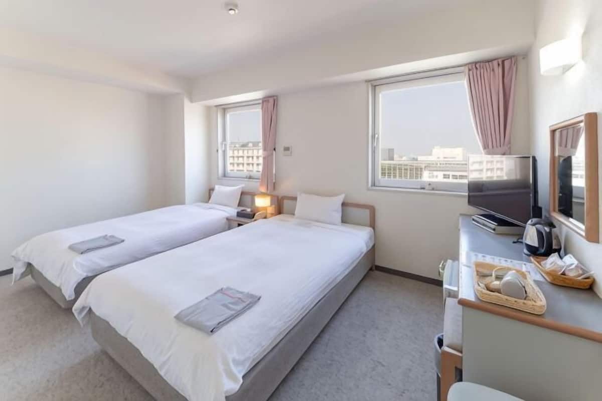Quarto de hotel com duas camas, uma escrivaninha com computador, televisão e janela com vista para a cidade.