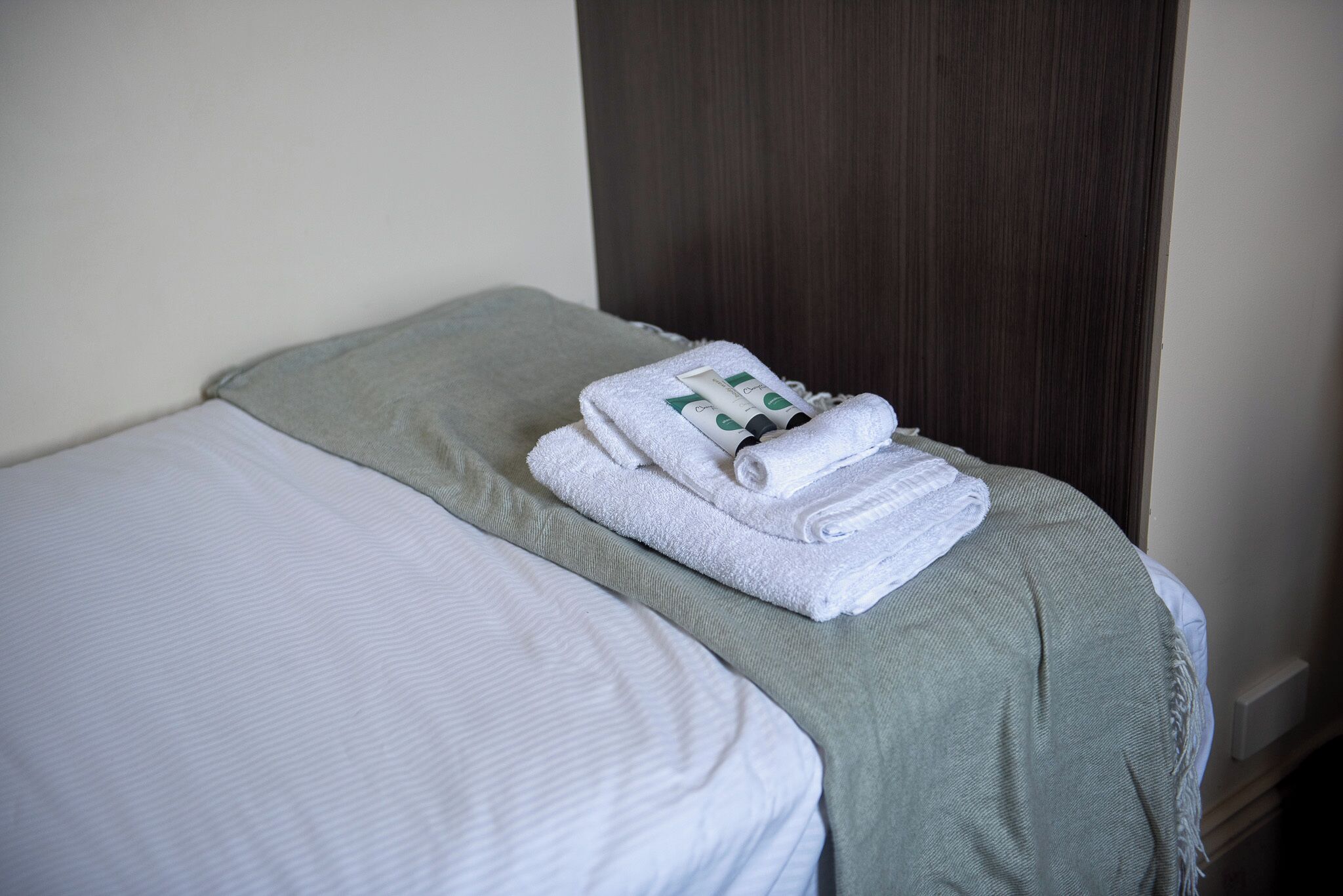 Cama bem arrumada com lençóis brancos e uma toalha dobrada sobre um cobre-leito verde.