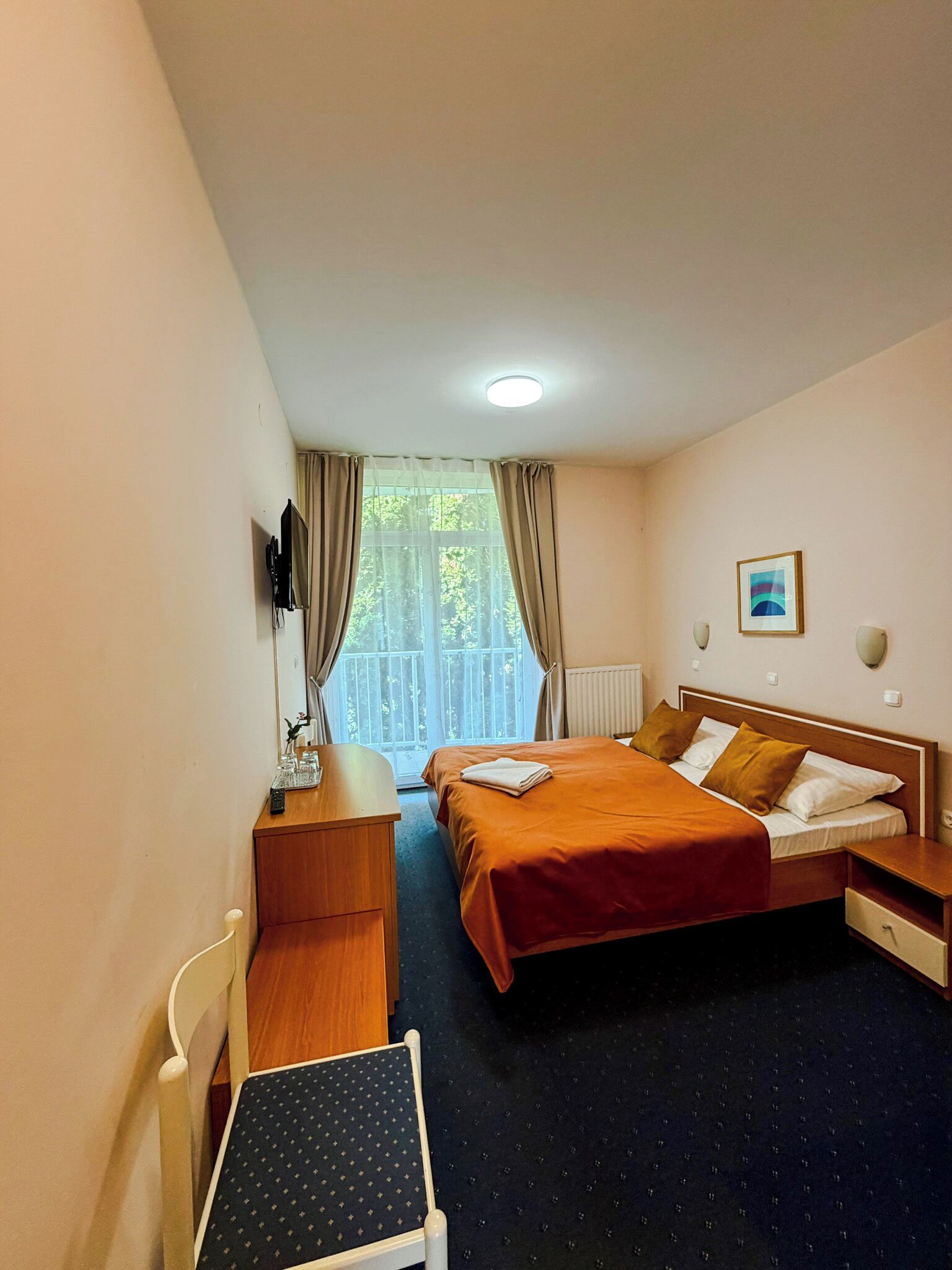 Quarto de hotel com cama, escrivaninha, cadeira, janela com cortinas, quadro na parede e radiador.