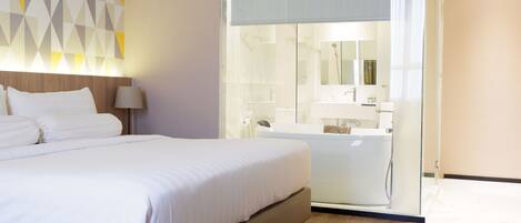 Quarto de hotel moderno com uma cama grande, mesa de cabeceira com abajur e banheiro visível por uma porta de vidro.