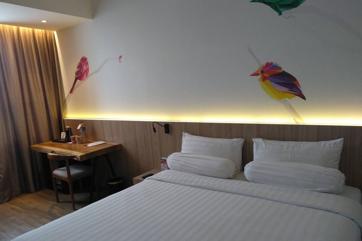 Quarto de hotel com cama, escrivaninha e cadeira. Há uma luminária na parede acima da escrivaninha.