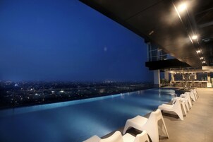Piscina infinita no terraço com espreguiçadeiras brancas, área de bar e vista da cidade ao anoitecer.