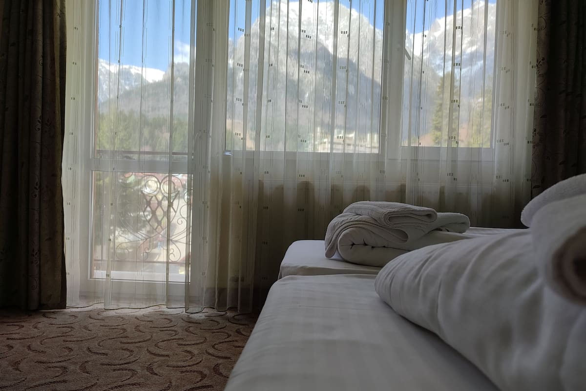 Quarto de hotel com cama, janela com cortinas leves e vista para montanhas.