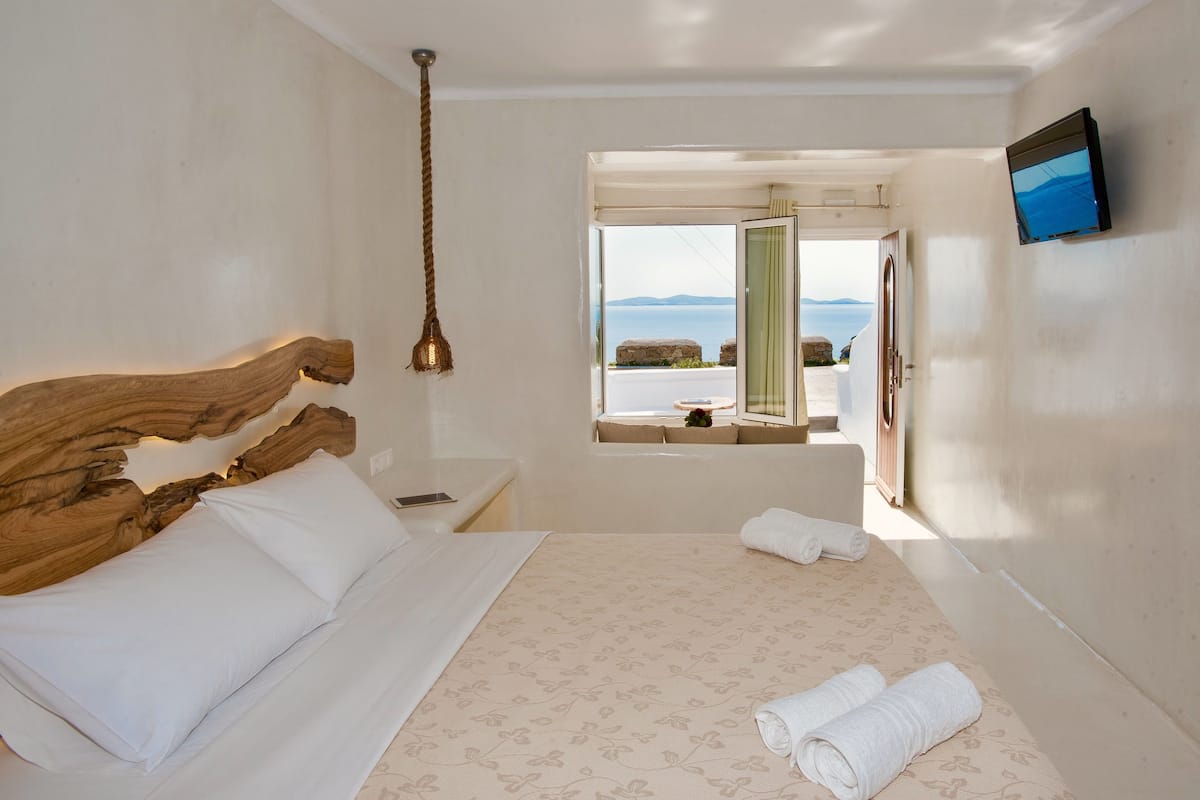 Um quarto com cama, cabeceira de madeira, mesa de cabeceira, luminária pendente, janela com vista para o mar e televisão na parede.