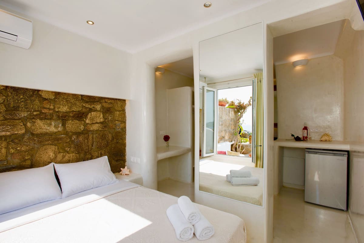 Quarto de hotel moderno com parede de destaque em pedra, uma cama grande com roupa de cama branca, um banheiro com espelho e uma cozinha compacta.