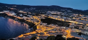 Uma cidade costeira ao entardecer, com ruas e edifícios iluminados, uma linha costeira visível e um cenário de colinas.