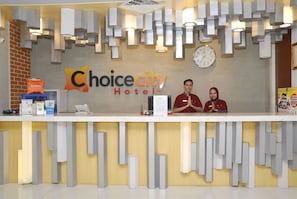 Bancada de recepção no Choice City Hotel, com relógio, mural de avisos e balcão com diversos objetos.