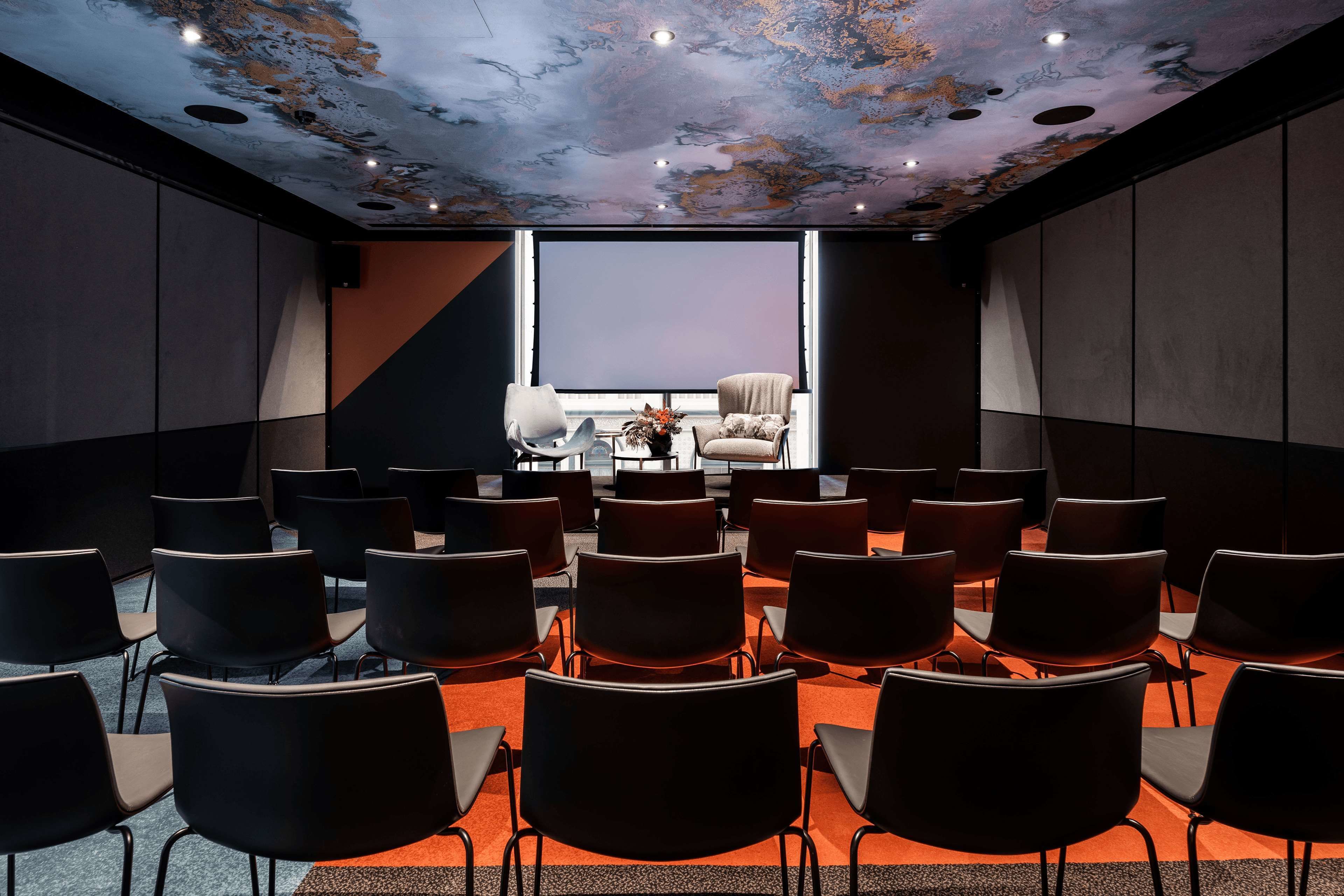 Uma sala de conferências com fileiras de cadeiras pretas, uma tela grande na frente e um teto com design exclusivo.