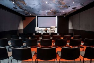 Uma sala de conferências com fileiras de cadeiras pretas, uma tela grande na frente e um teto com design exclusivo.