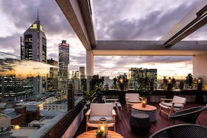 Terraço no telhado com área de estar e vista para a cidade ao entardecer.