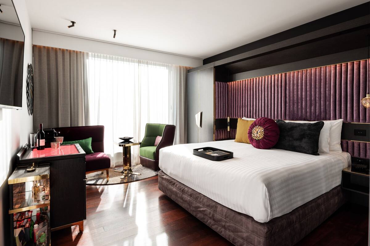 Quarto de hotel moderno com uma cama grande, escrivaninha com minibar, poltrona verde e vista para a cidade.