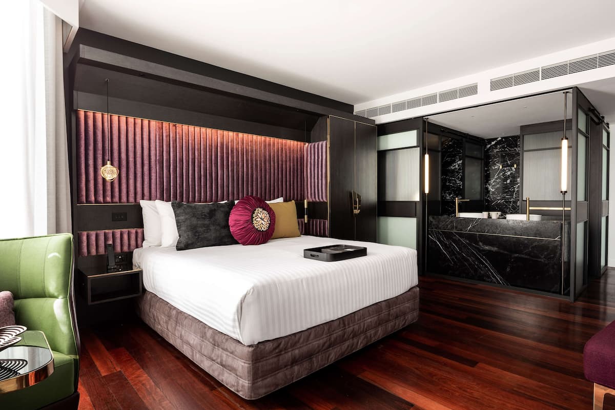 Quarto de hotel moderno com uma cama grande, uma poltrona verde, uma mesinha e um banheiro com bancada de mármore.