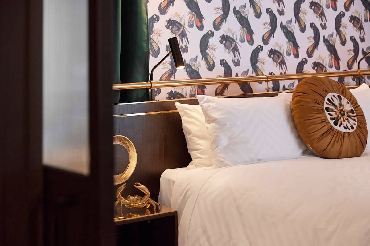 Uma cama bem arrumada com roupa de cama branca e um travesseiro decorativo, uma cabeceira com moldura dourada e uma parede com padrão de pássaros.