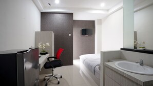Quarto de hotel moderno com cama, escrivaninha com computador, cadeira, banheiro com pia e espelho, e televisão fixada na parede.