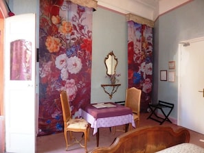 Quarto com cama, duas cadeiras, uma mesa com toalha de mesa roxa, espelho e cortinas florais.