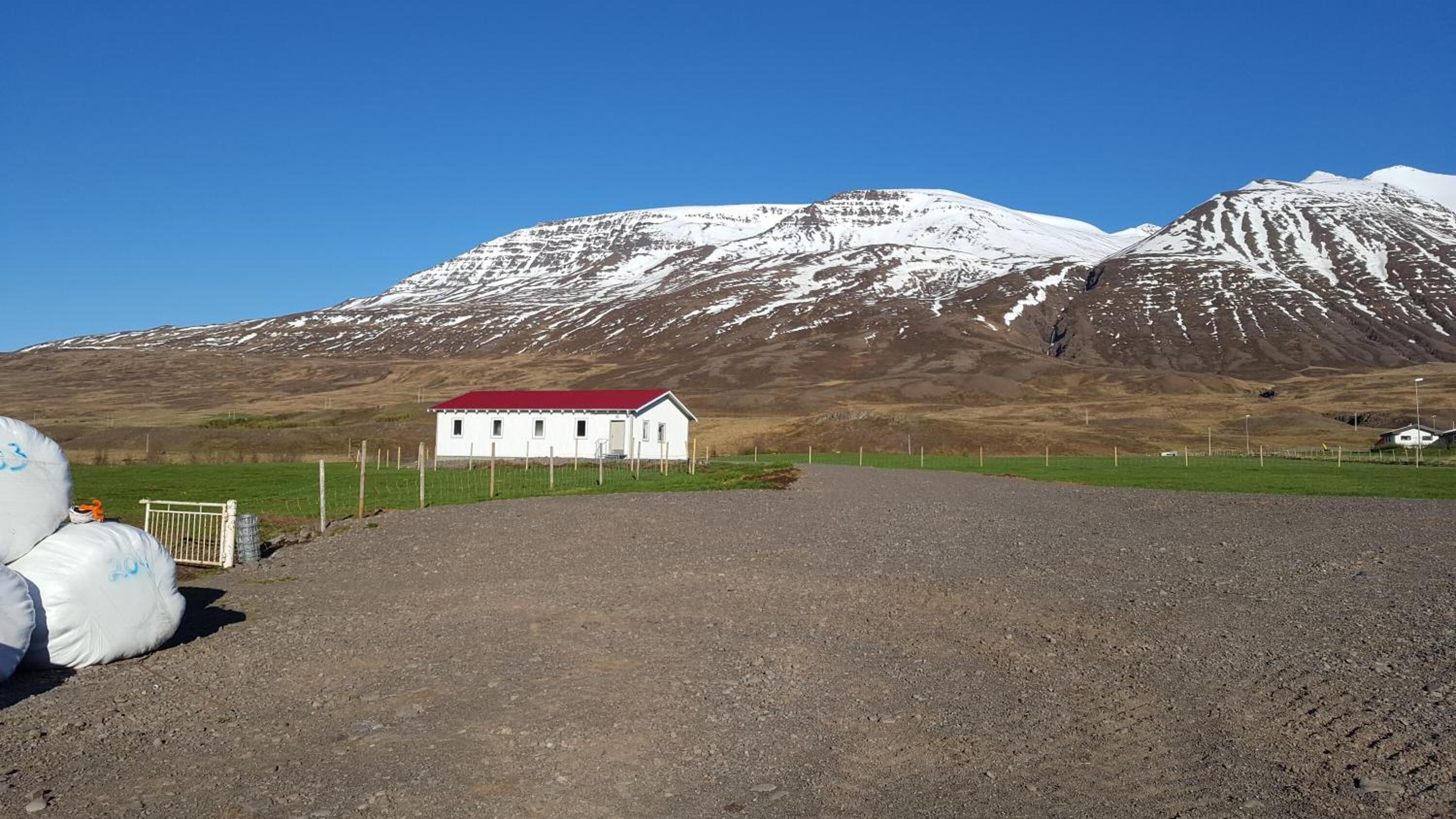 Um edifício branco com telhado vermelho, rodeado por um espaço com brita e uma cerca, com montanhas cobertas de neve ao fundo.
