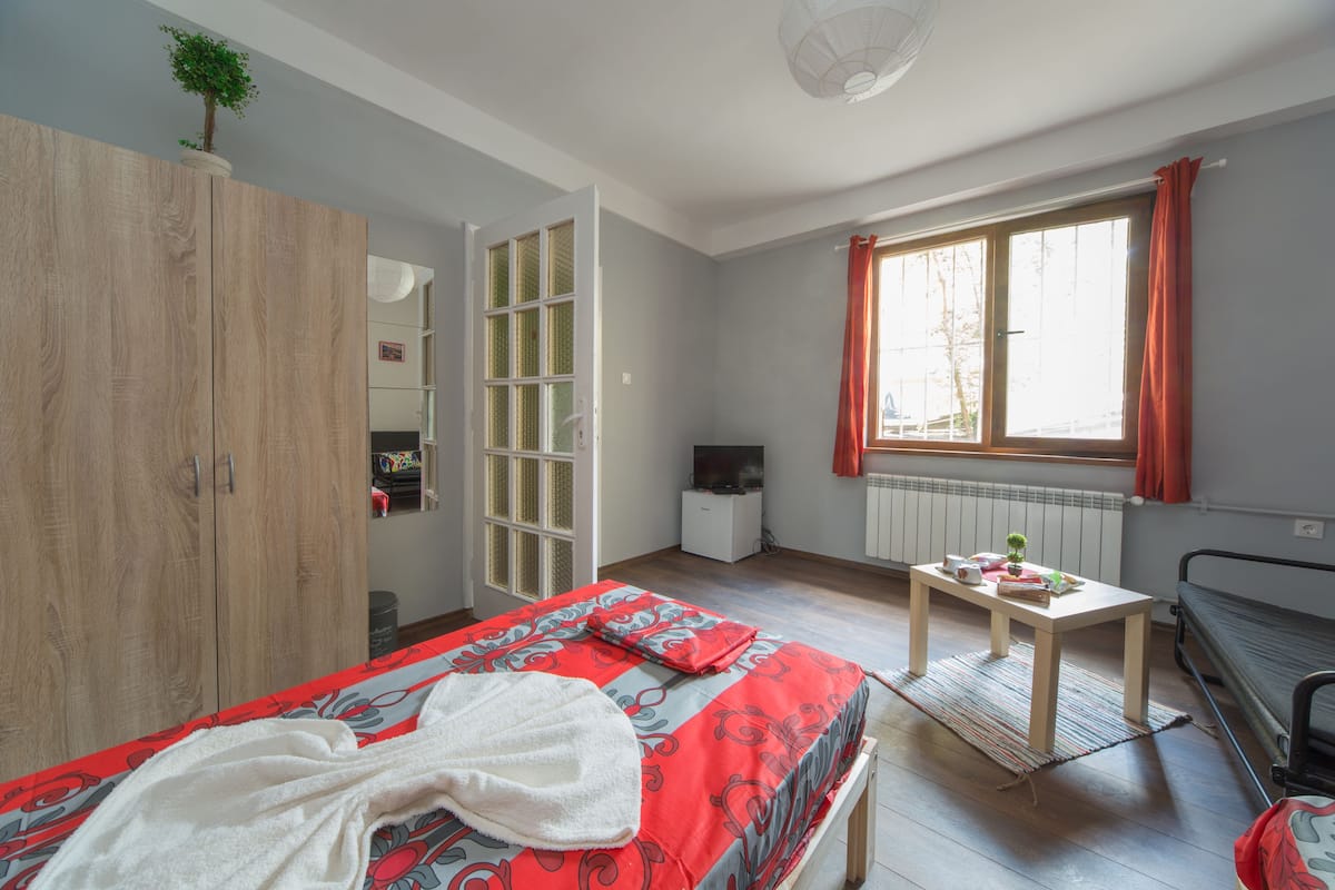 Quarto com cama, guarda-roupa de madeira, mesinha com planta, radiador e janela com cortinas.