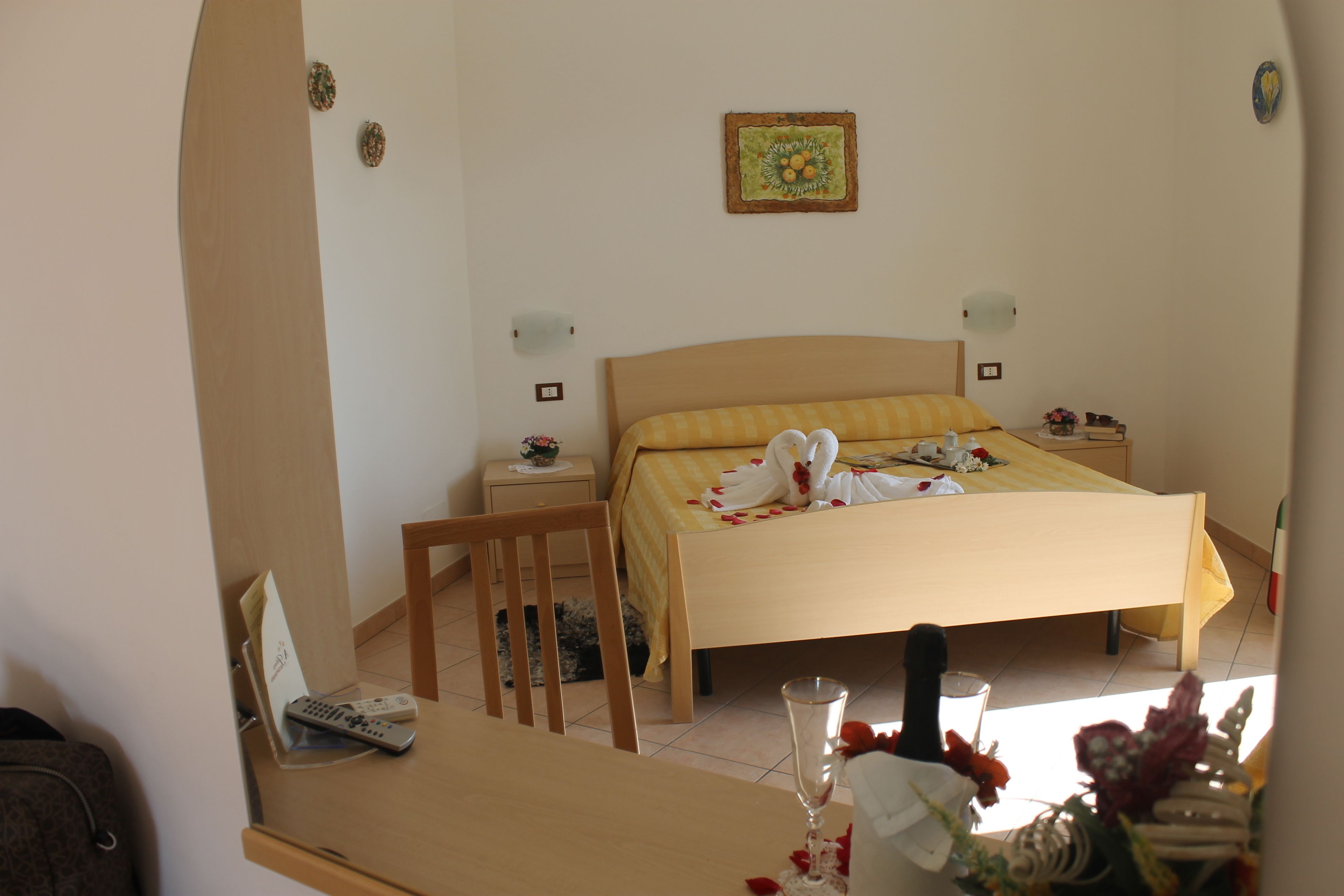 Quarto de hotel com uma cama, uma cadeira de madeira, uma mesa com um laptop, um copo de água e um vaso com flores.