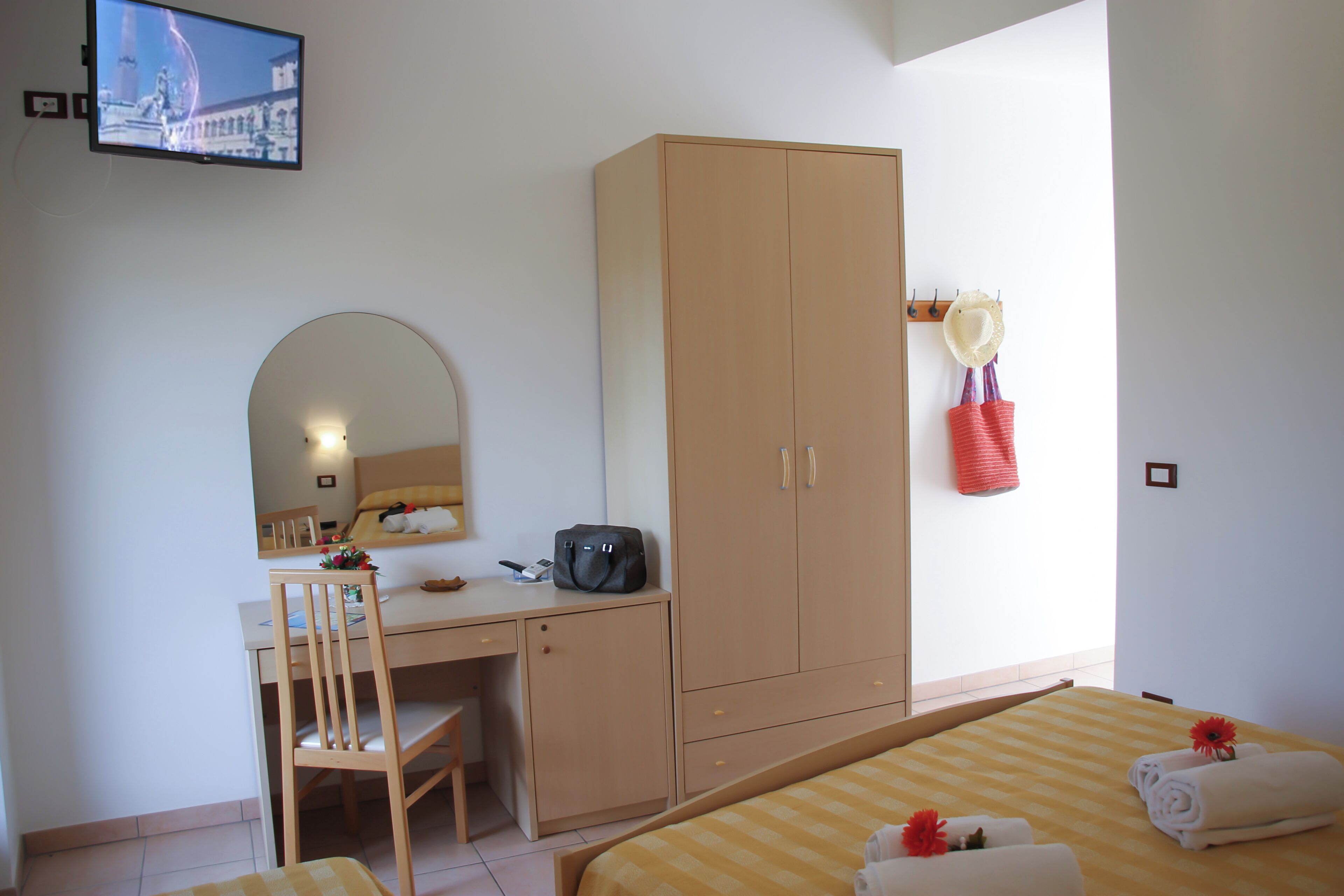 Um quarto com cama, escrivaninha, cadeira, guarda-roupa e televisão na parede.