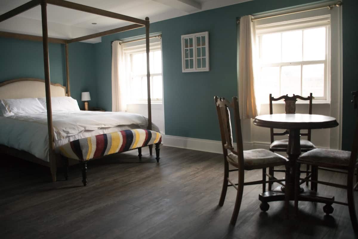 Uma cama com dossel e um painel de cabeceira listrado em cores vibrantes, uma mesa redonda de madeira com quatro cadeiras e uma janela com cortinas brancas.