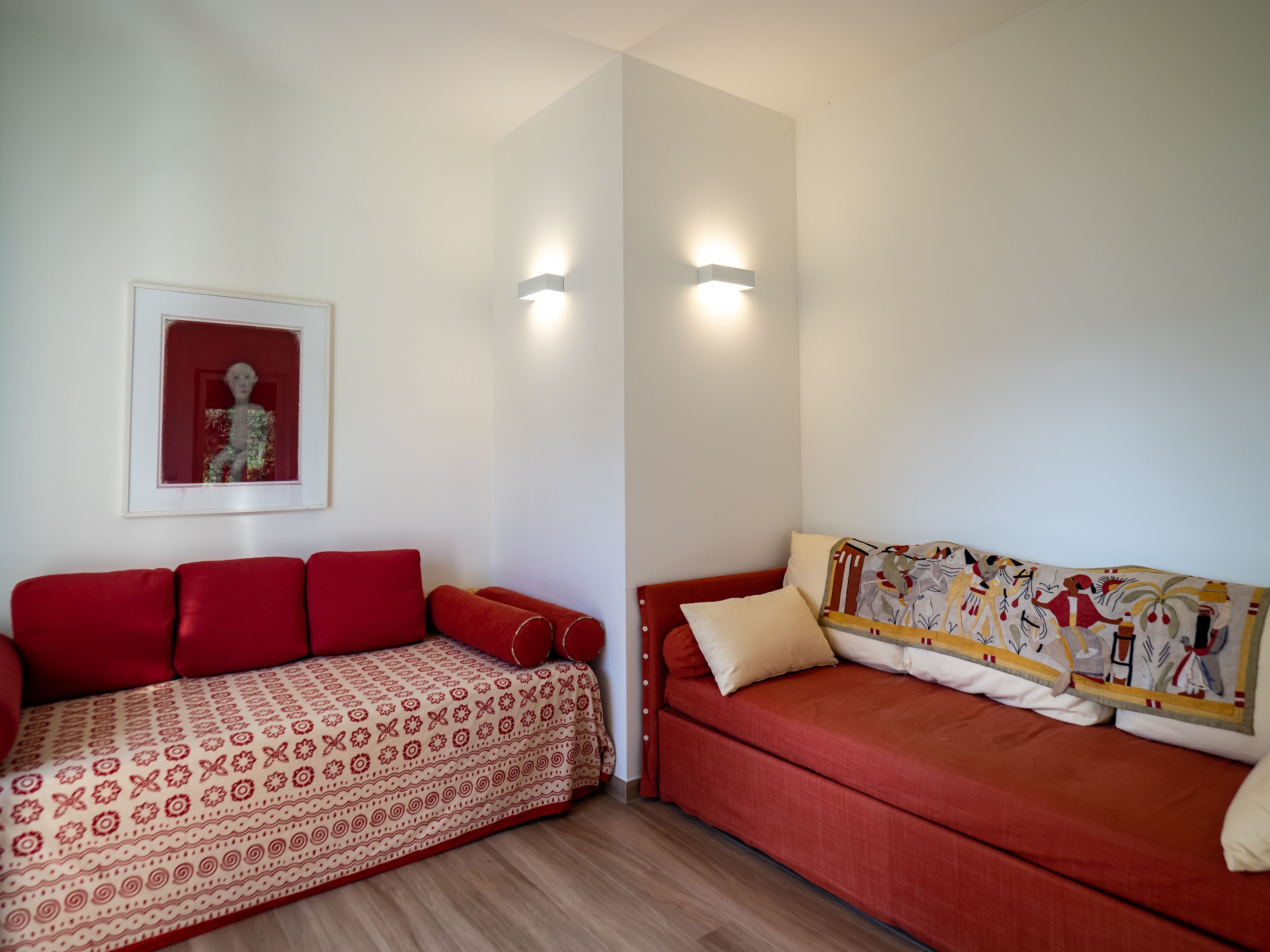 Quarto com um sofá vermelho e um sofá estampado, ambos com almofadas. Há um quadro na parede e duas luminárias fixadas.
