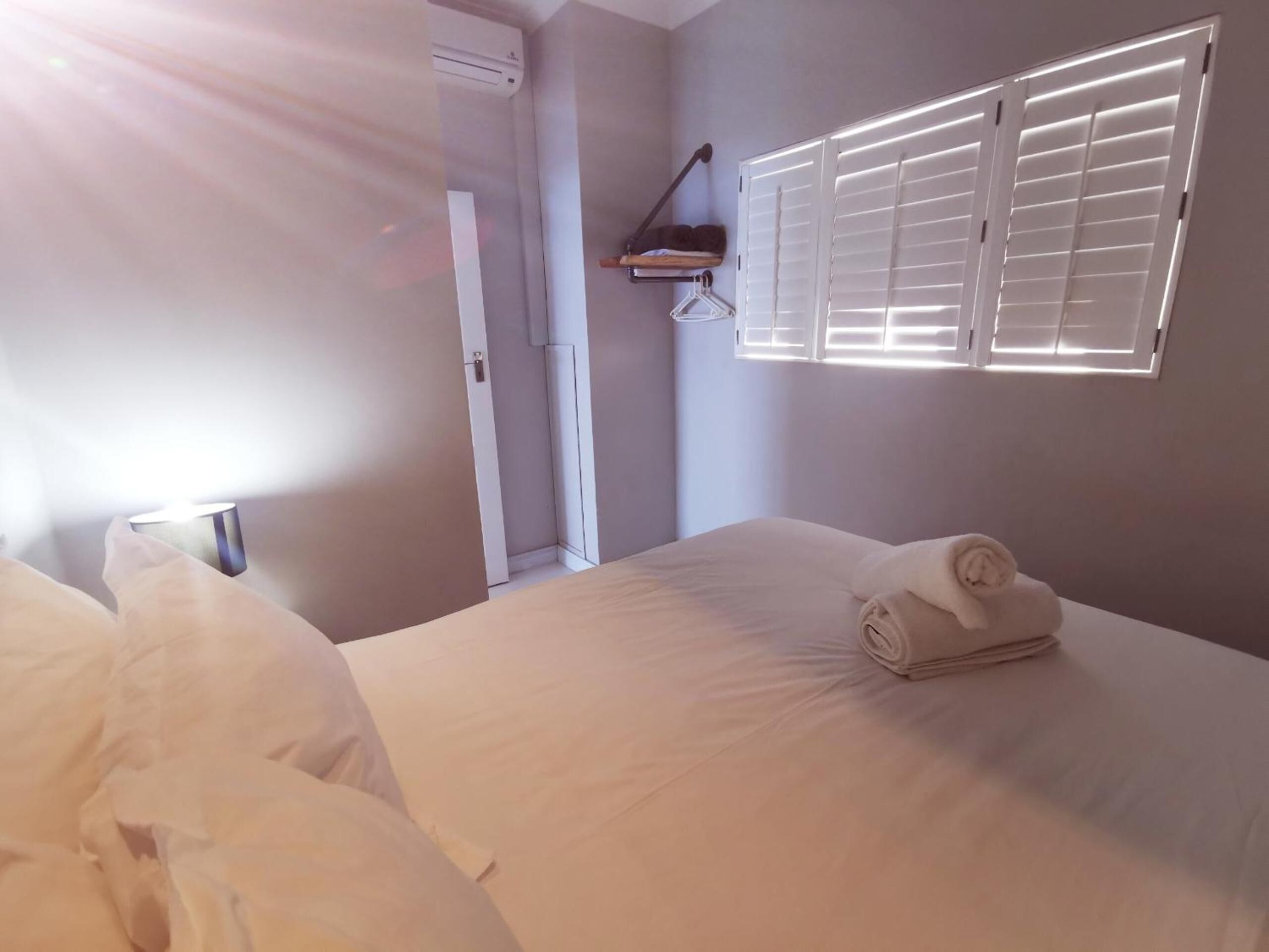Quarto de hotel com uma cama, mesa de cabeceira, luminária, janela com persianas e porta.