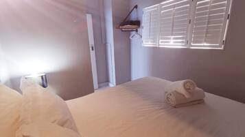 Quarto de hotel com uma cama, mesa de cabeceira, luminária, janela com persianas e porta.