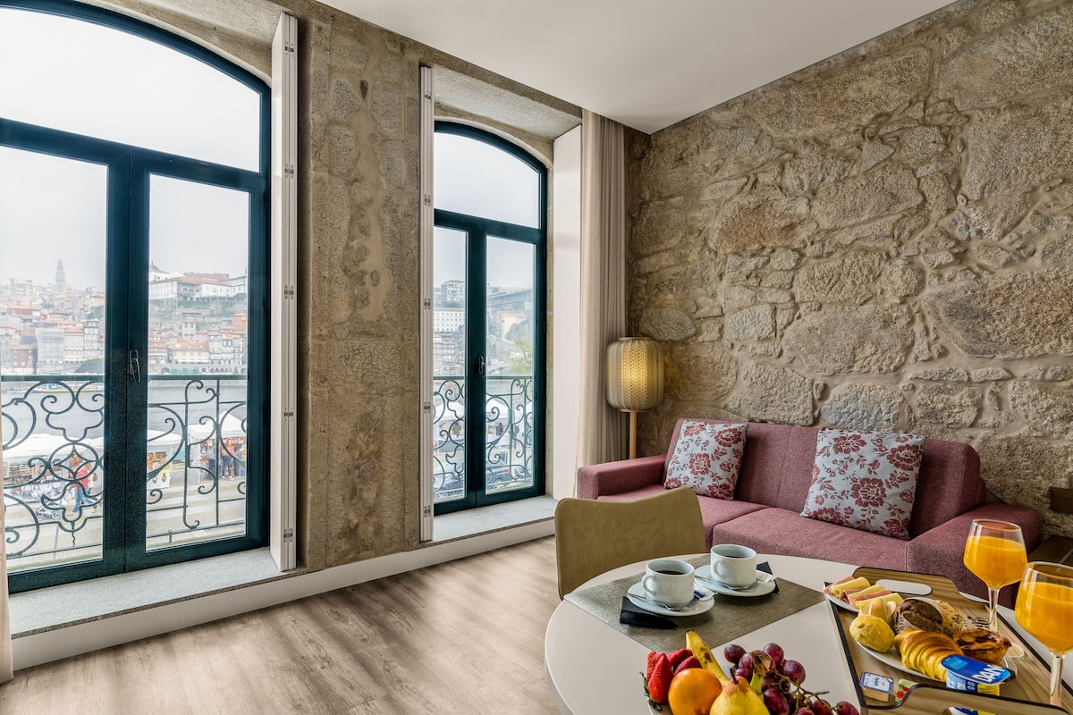 Quarto com parede de pedra, sofá, mesa de jantar com café da manhã e grandes janelas com vista para a cidade.